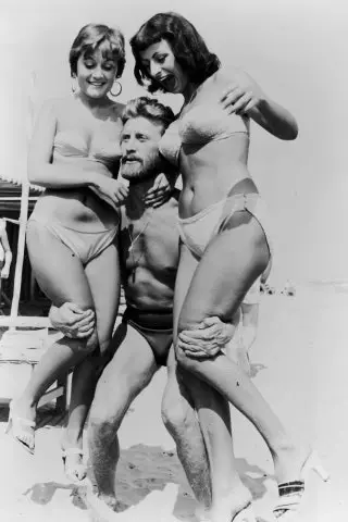 Kirk Douglas lig twee vroue wat bikini's dra op 'n strand tydens die Venesiese rolprentfees 1953